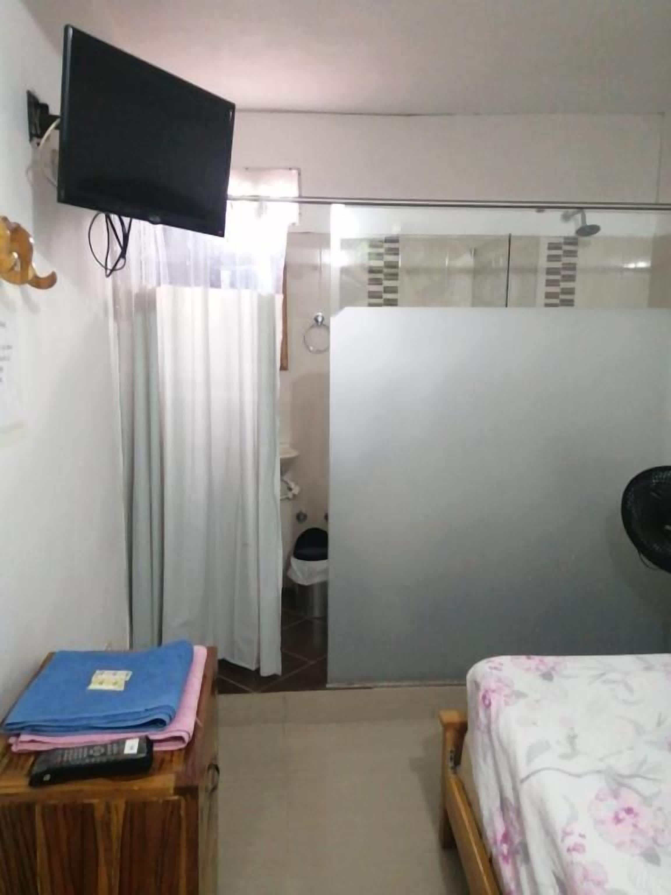 Quarto com cama, televisão na parede, área de chuveiro e uma pequena mesa com objetos.
