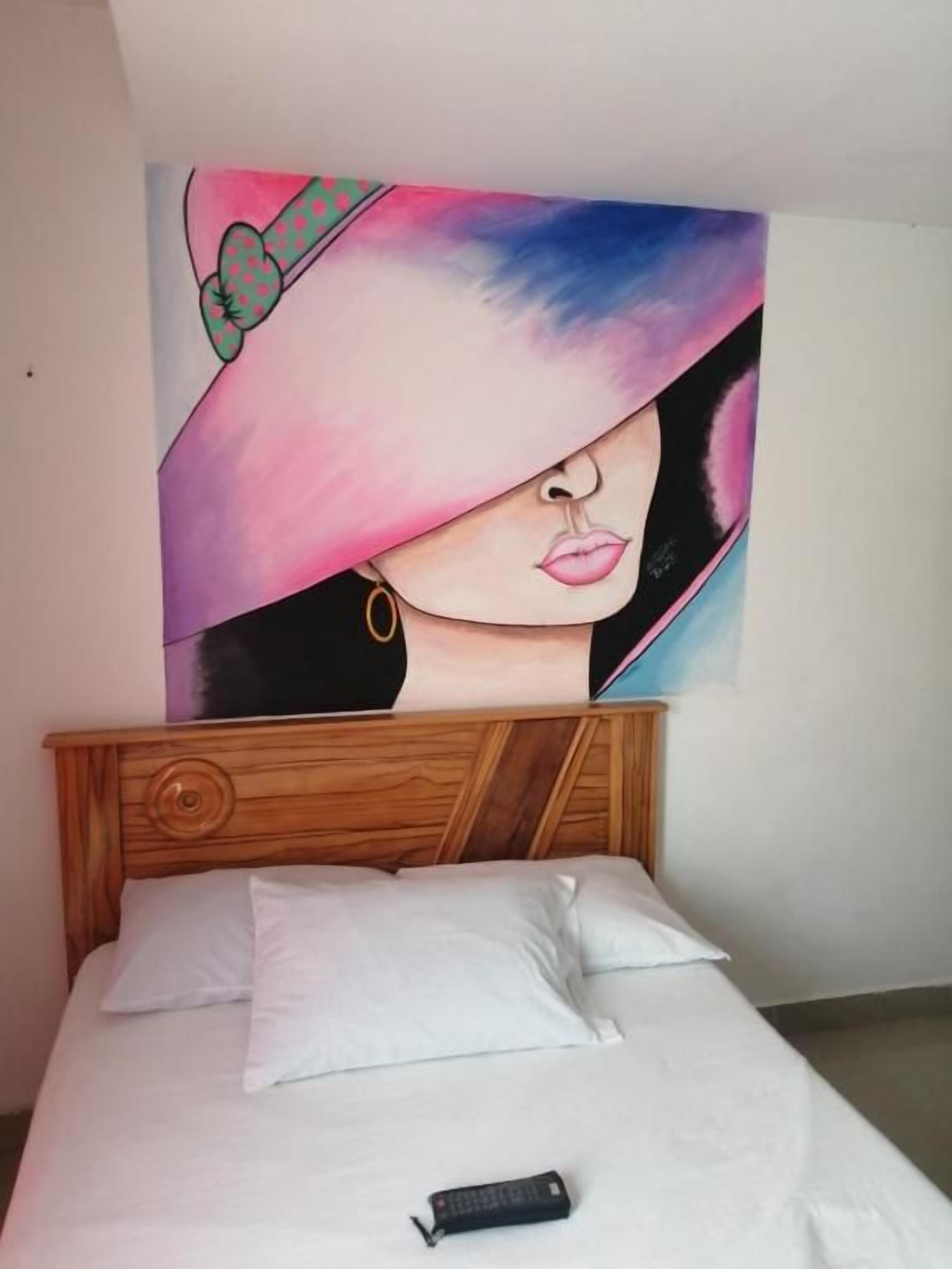 Cama com lençóis brancos, um controle remoto sobre a cama e uma grande pintura abstrata acima.