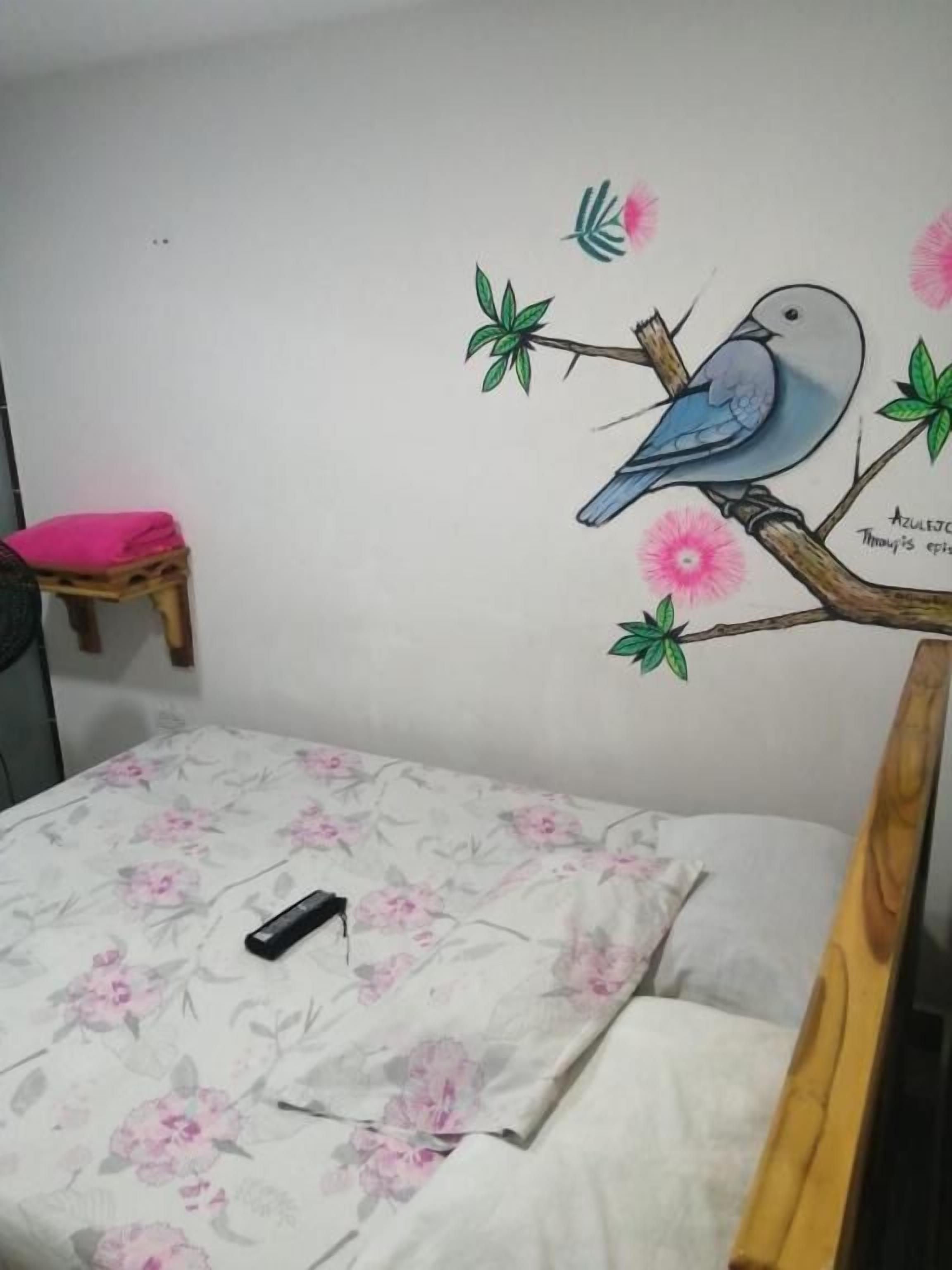 Quarto com uma cama, um mural de um pássaro na parede e um almofada rosa em uma cadeira.