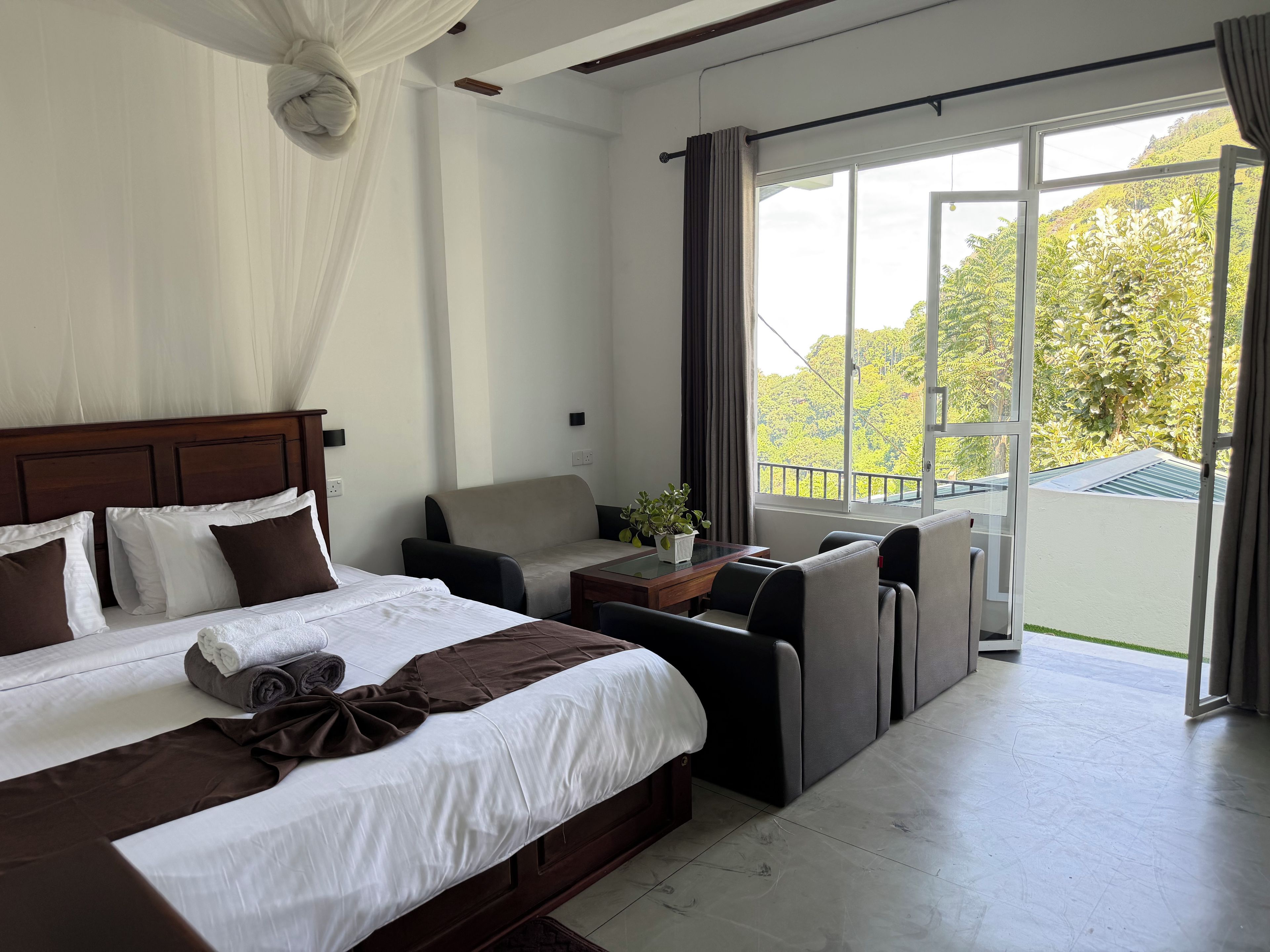Quarto de hotel com uma cama grande, um sofá, uma mesinha com uma planta e uma varanda com vista para o verde.