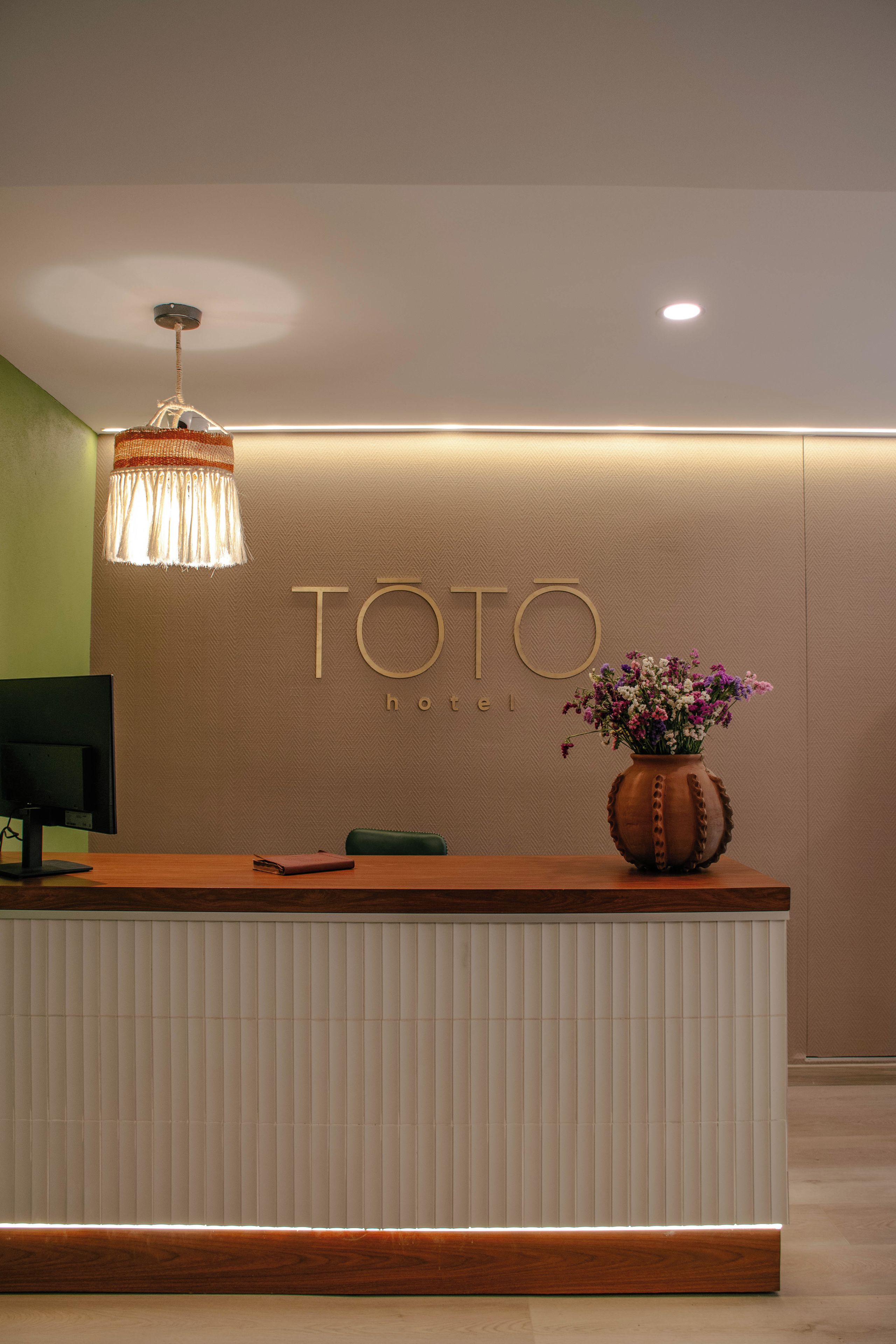 Uma receção com design moderno, um monitor, um vaso com flores e uma parede com o nome 'TOTO HOTEL' em exposição.