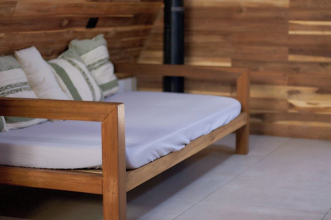 Uma cama de madeira com colchão e almofadas brancas, diante de uma parede de madeira e piso de azulejos.