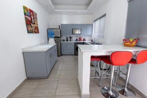 Uma cozinha compacta com armários cinzentos, bancada branca e uma pequena ilha com banquetas vermelhas.