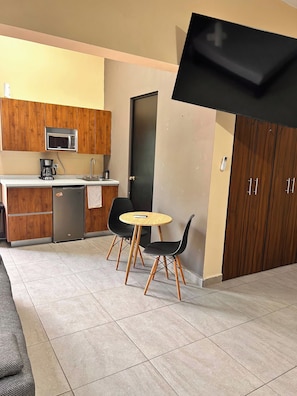 Cozinha compacta com micro-ondas, cafeteira e pequena geladeira. Há uma área de refeições com duas cadeiras e uma mesa redonda pequena.