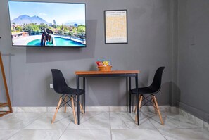 Quarto com televisão na parede exibindo uma vista panorâmica, uma obra de arte emoldurada acima da TV, e uma mesa com duas cadeiras e um cesto de lanches.