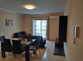Uma sala de estar moderna com área de jantar, um conjunto de sofás, uma TV de tela plana e uma grande janela com cortinas.