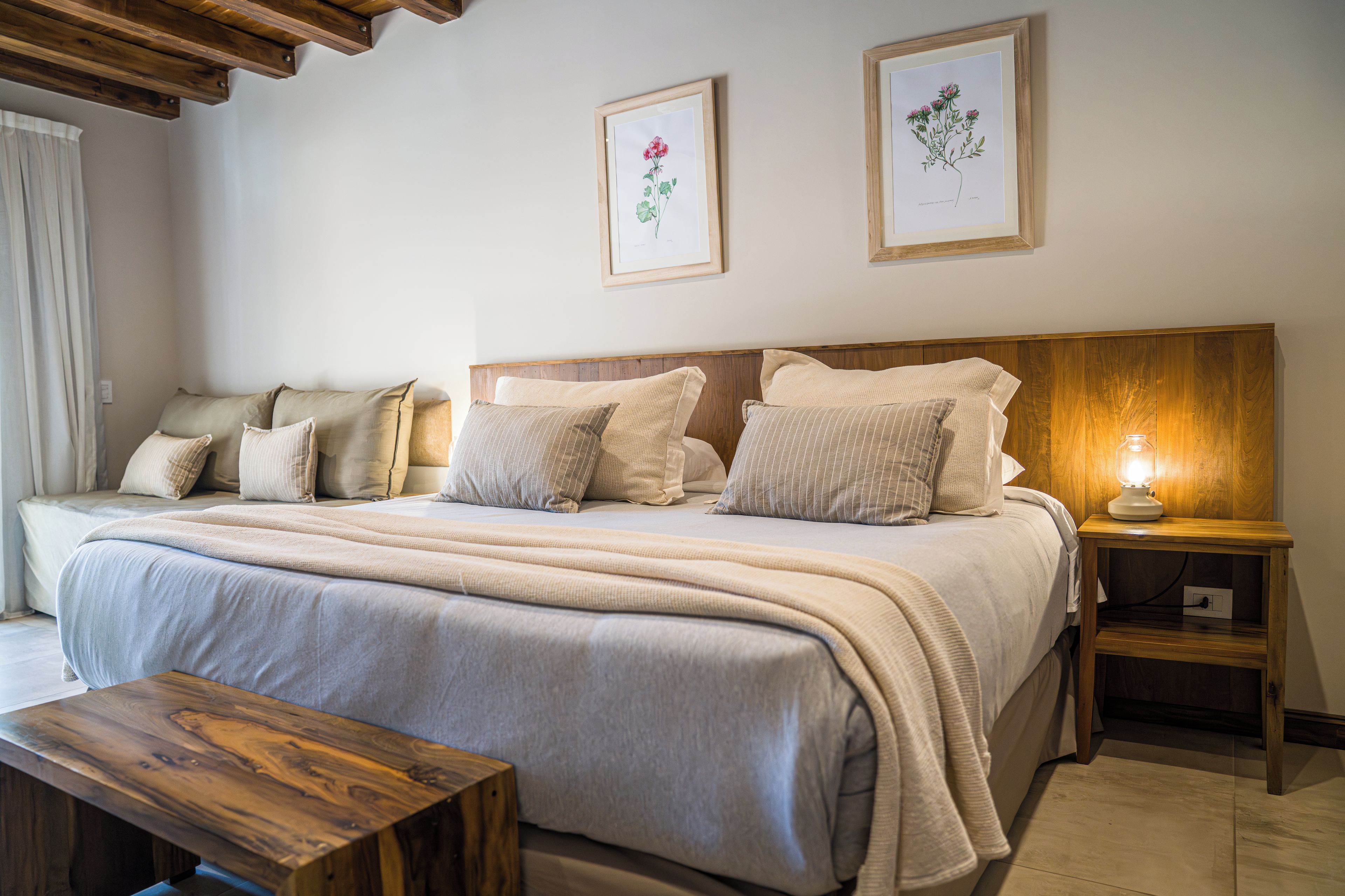 Quarto com cabeceira de madeira, cama com várias almofadas, mesa de cabeceira com abajur e duas ilustrações botânicas emolduradas na parede.