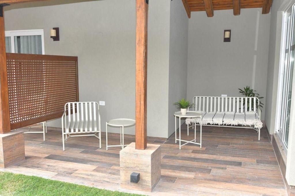 Uma área de varanda coberta com pérgola de madeira, duas cadeiras brancas, uma pequena mesa redonda e um sofá branco.