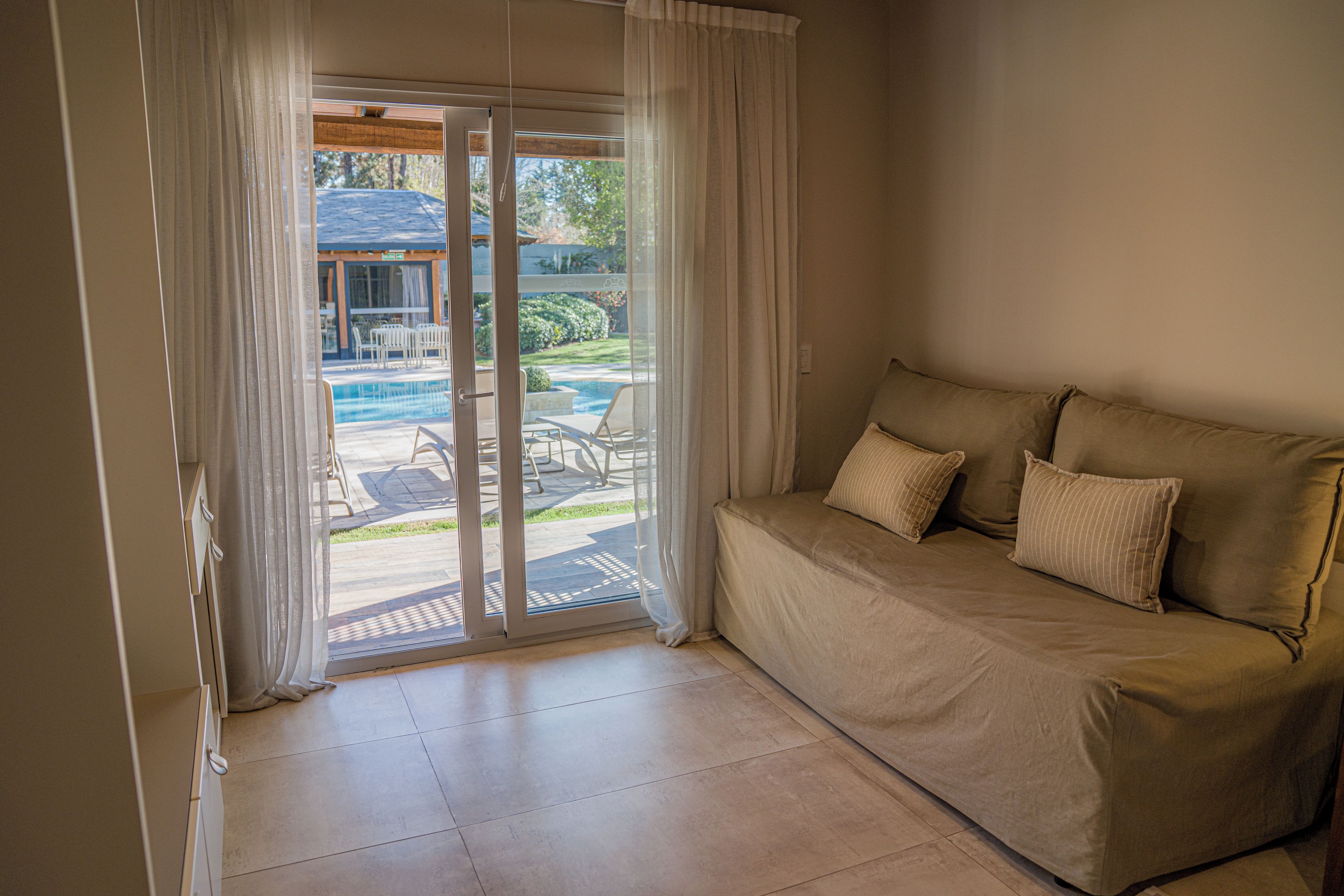 Quarto com porta de vidro deslizante para área da piscina, sofá bege com almofadas e piso ladrilhado.