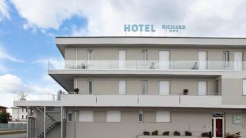 Edifício de hotel de dois andares com fachada plana, varanda no andar superior e porta vermelha na entrada.