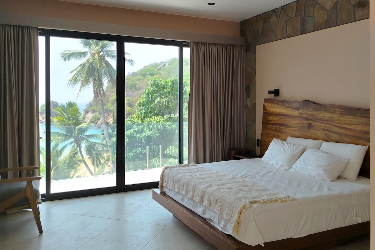 Quarto de hotel com cama grande, parede decorada com pedra e vista para palmeiras e praia.