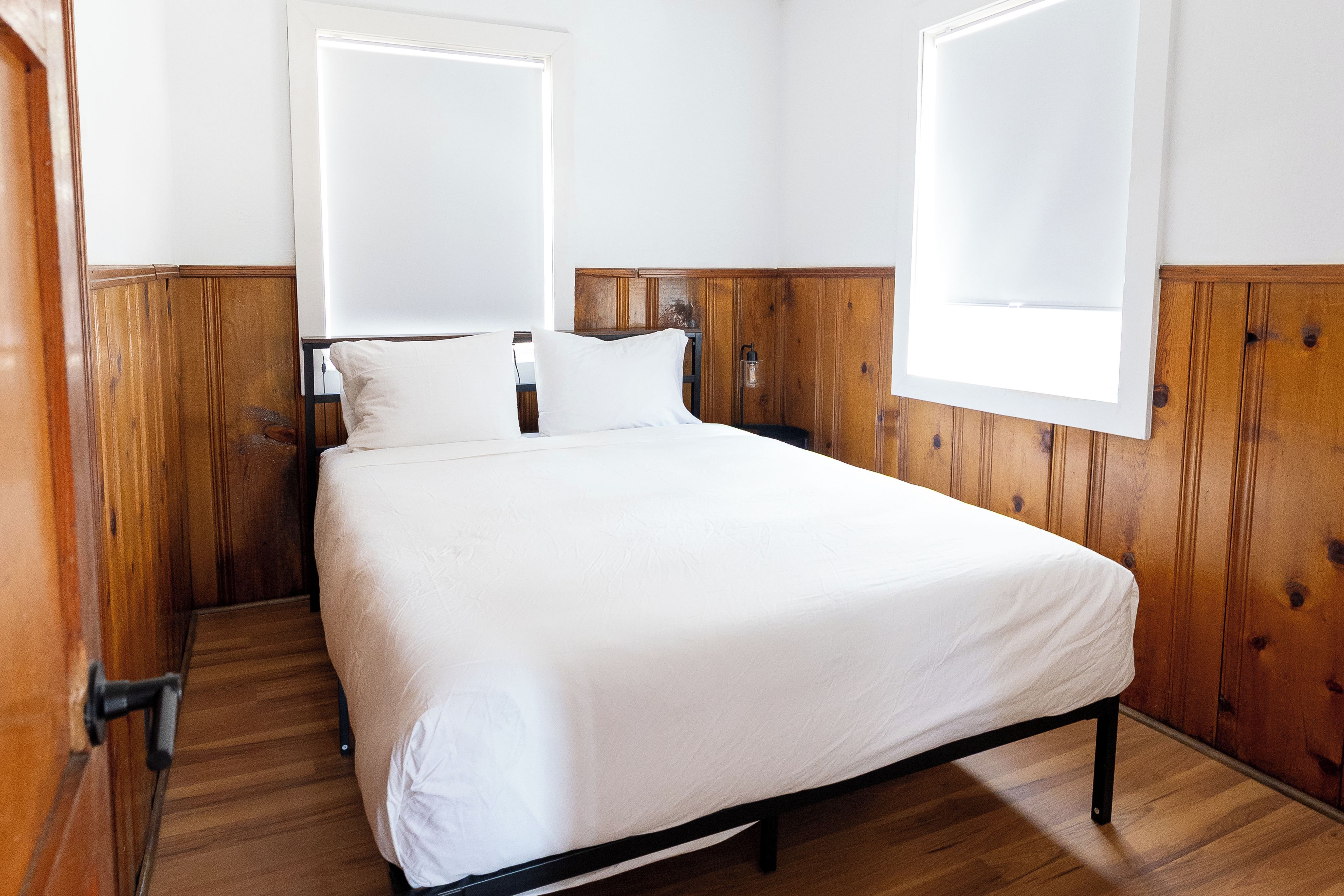 Cama de solteiro com cabeceira e peseira de madeira, roupa de cama branca e janela com persianas.