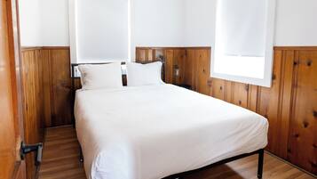 Cama de solteiro com cabeceira e peseira de madeira, roupa de cama branca e janela com persianas.