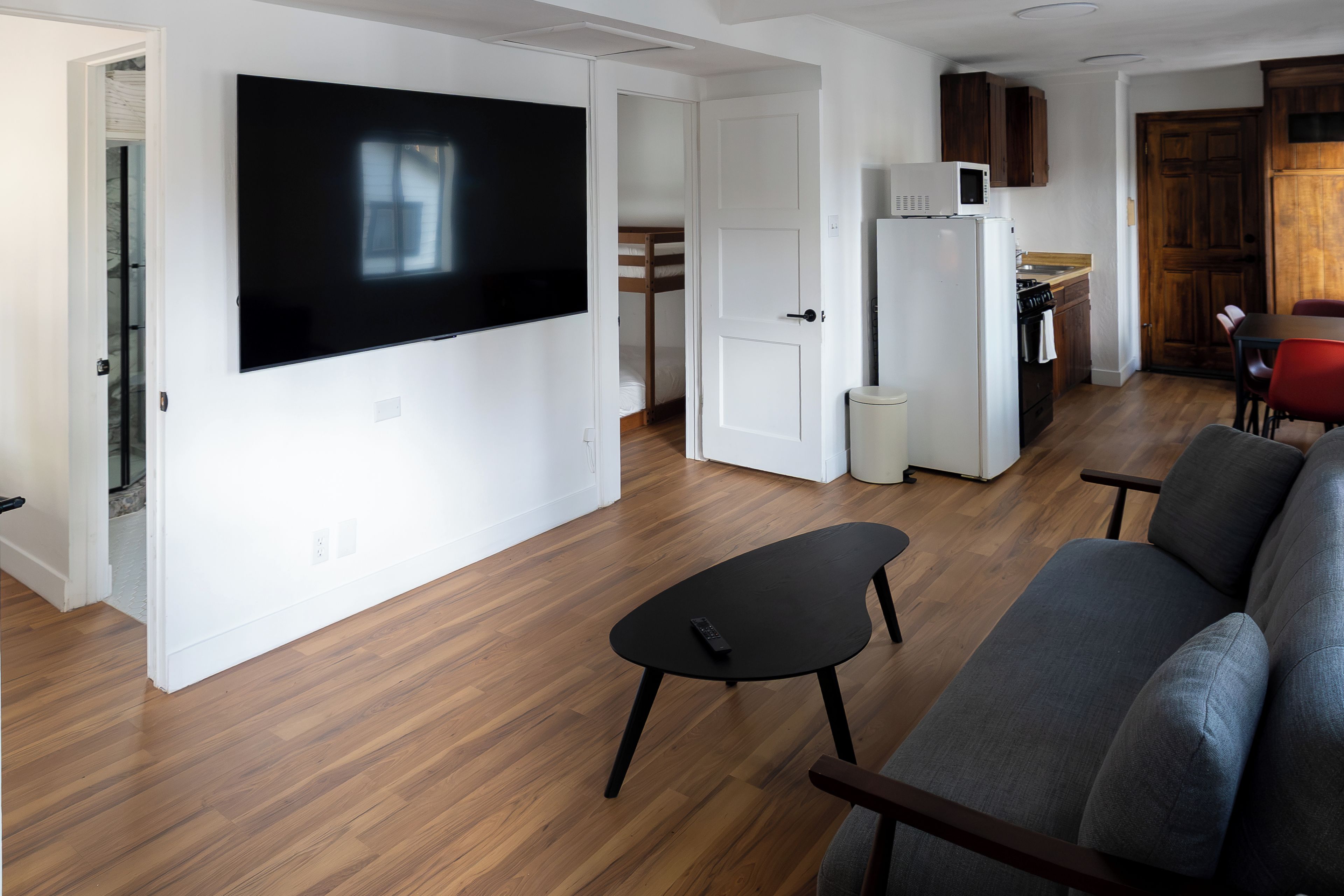 Uma sala de estar moderna com televisão de tela plana na parede, um sofá, uma mesinha de centro preta e uma área de cozinha com micro-ondas e geladeira.