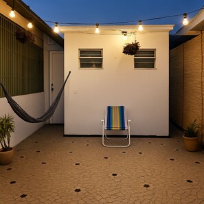 Quarto com rede, cadeira listrada e parede com duas janelas e plantas penduradas.