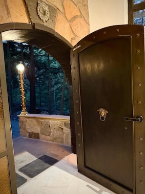 Uma porta de madeira escura com maçaneta de metal e fechadura, dando acesso a uma área externa com um banco de pedra e um poste de luz.