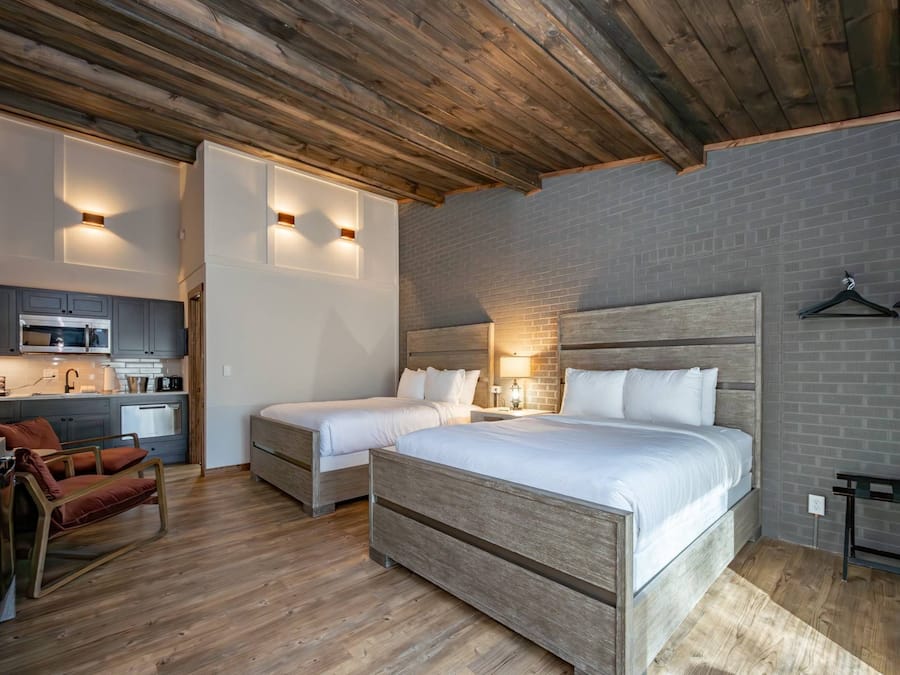 Quarto de hotel com teto de madeira, parede de tijolos, cama com roupa de cama branca, pequena cozinha e área de estar.