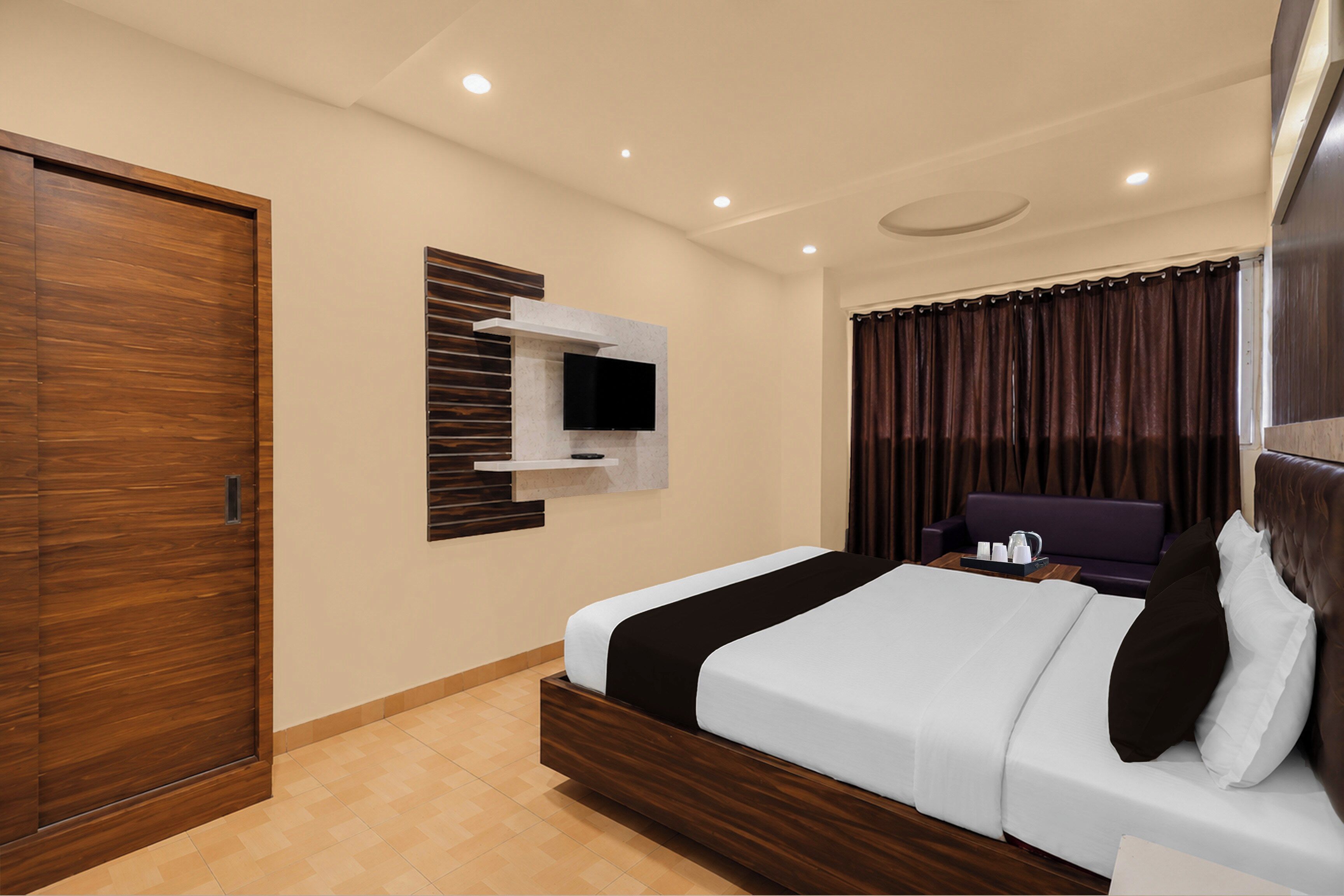 Quarto de hotel moderno com guarda-roupa de madeira, cama com colcha preta e branca, televisão montada e sofá de cor escura.