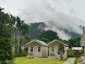 Uma casa de dois andares com telhado plano, varanda frontal com cadeiras e vista para uma paisagem montanhosa.