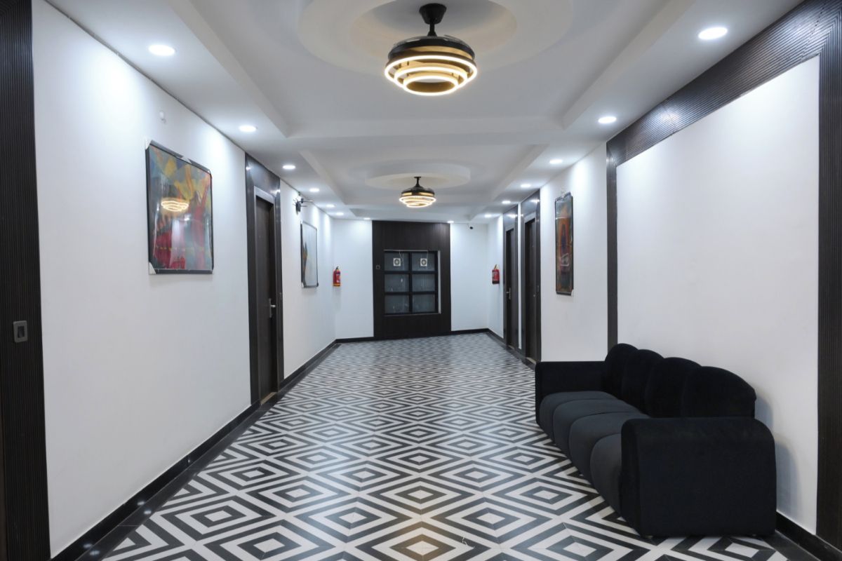 Corredor de hotel com piso estampado, área de estar com cadeiras estofadas pretas e luminária no teto.