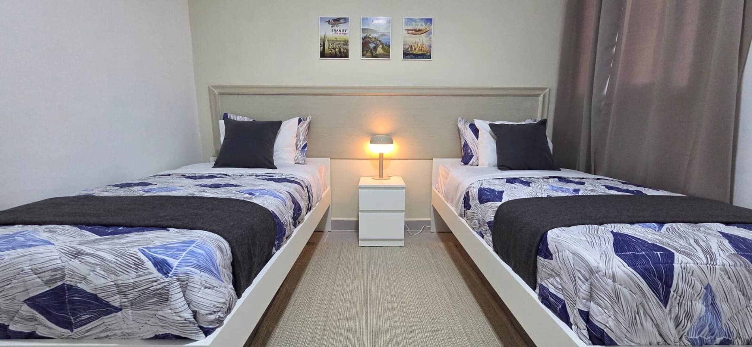 Quarto de hotel com duas camas de solteiro, mesa de cabeceira com abajur e três quadros na parede.