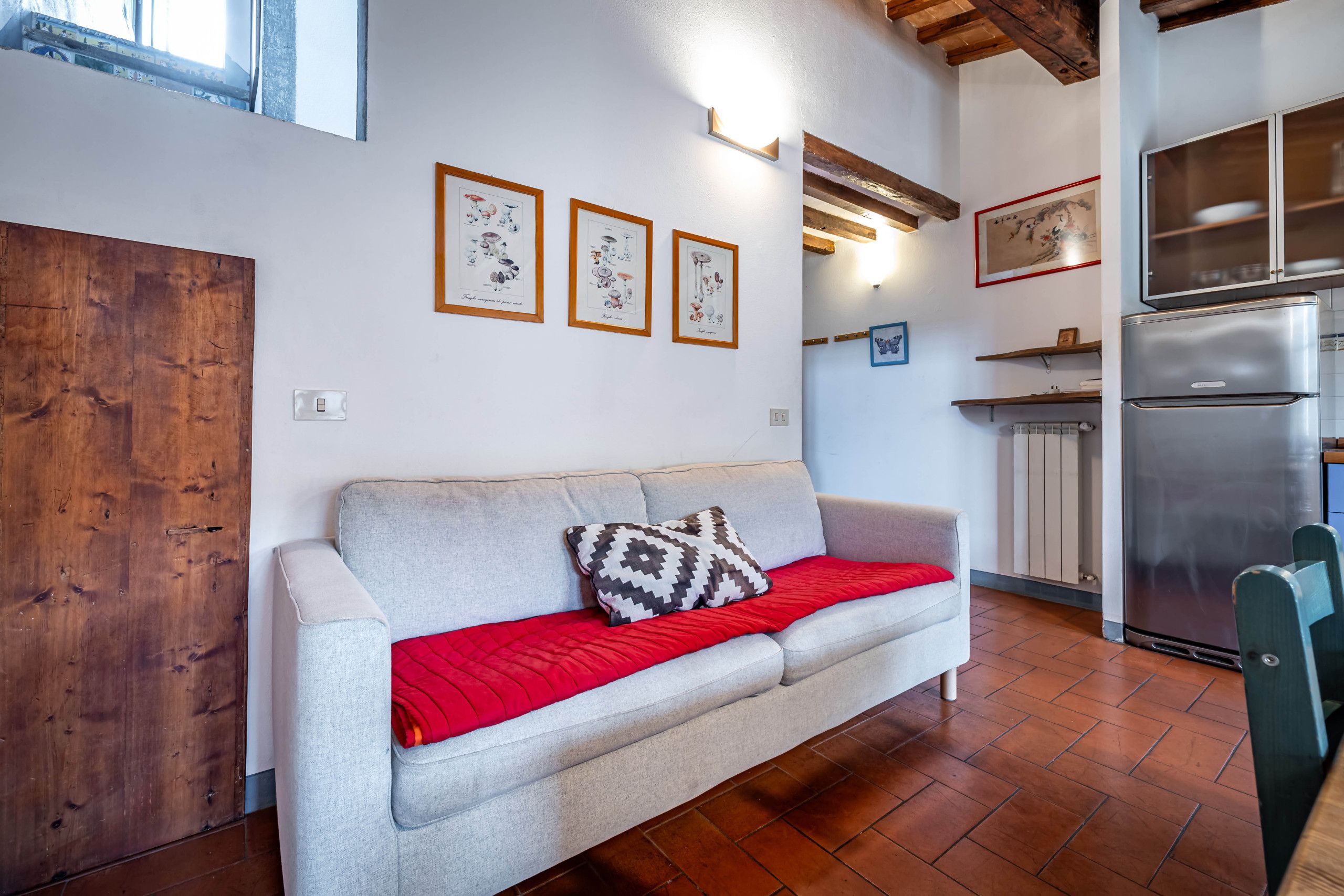 Sala de estar com sofá, manta vermelha e porta de madeira. Há quadros nas paredes e uma área de cozinha com refrigerador.