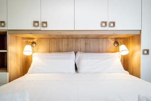 Cama com duas almofadas, cabeceira de madeira e duas luminárias fixadas na parede.
