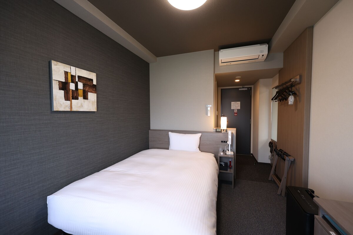 Quarto de hotel com cama, mesa de cabeceira, aparelho de ar condicionado, porta e corredor.