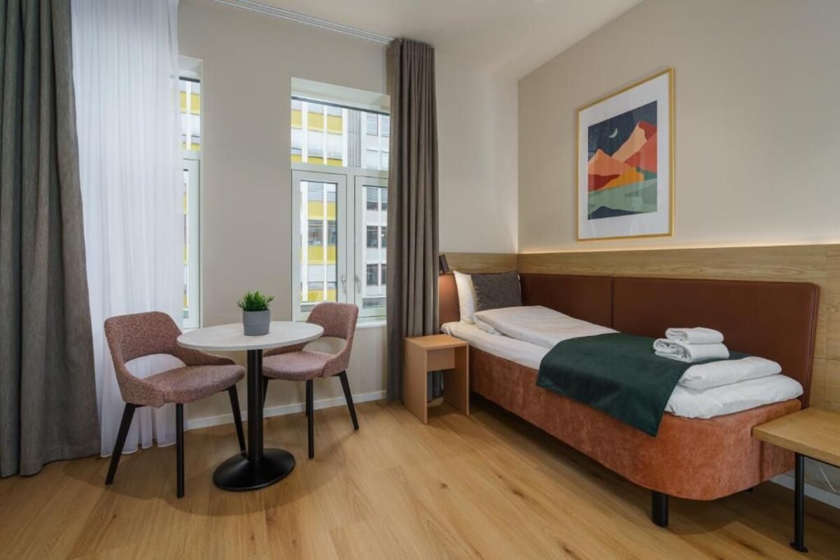 Quarto de hotel com cama, pequena mesa com duas cadeiras, mesa de cabeceira, janela com cortinas e um quadro na parede.