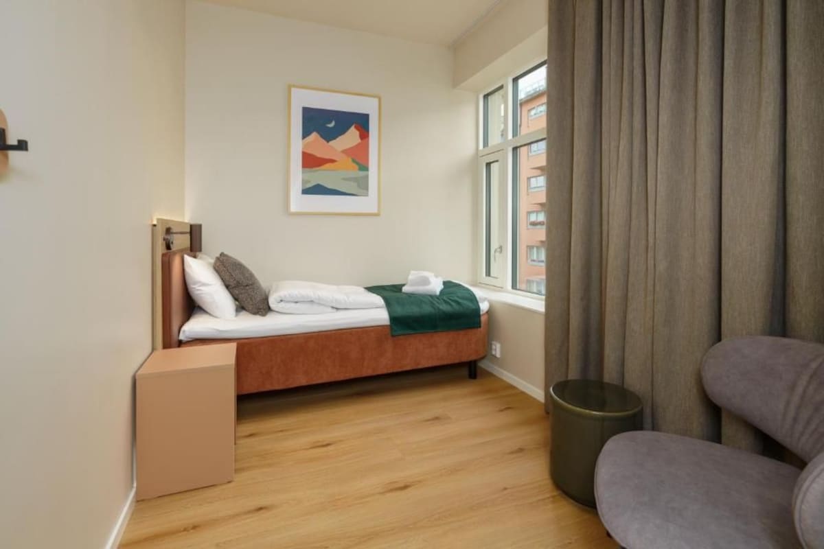 Quarto com uma cama de solteiro, janela com cortinas, uma pequena lixeira e um quadro na parede.