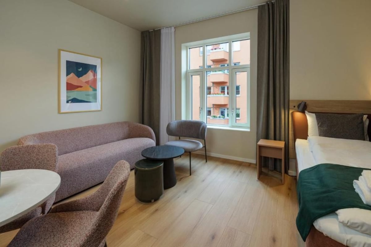 Quarto de hotel com cama, sofá, mesa pequena e cadeira. Janela com vista para edifícios.