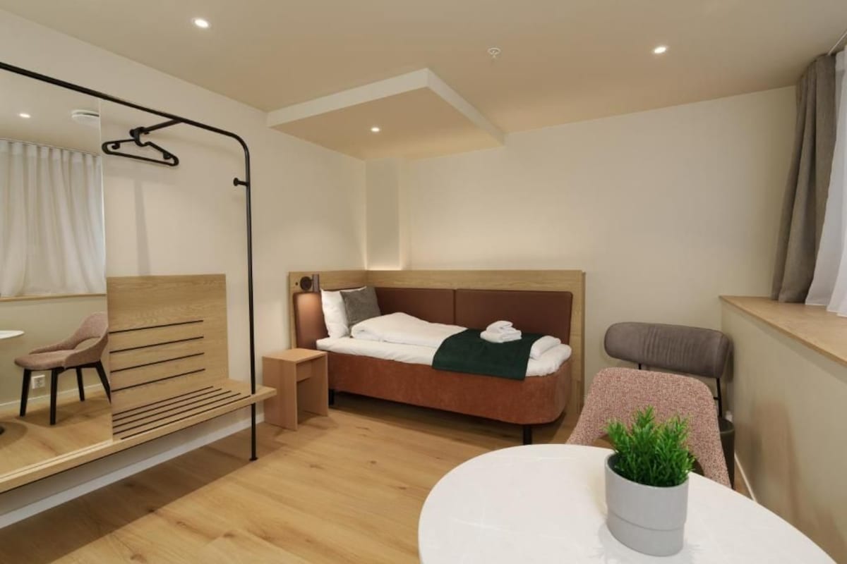 Quarto de hotel moderno com cama, mesa de cabeceira, cadeira e uma pequena mesa com uma planta.