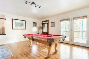 Mesa de bilhar em um espaço com piso de madeira, quadro na parede e janela com persianas.