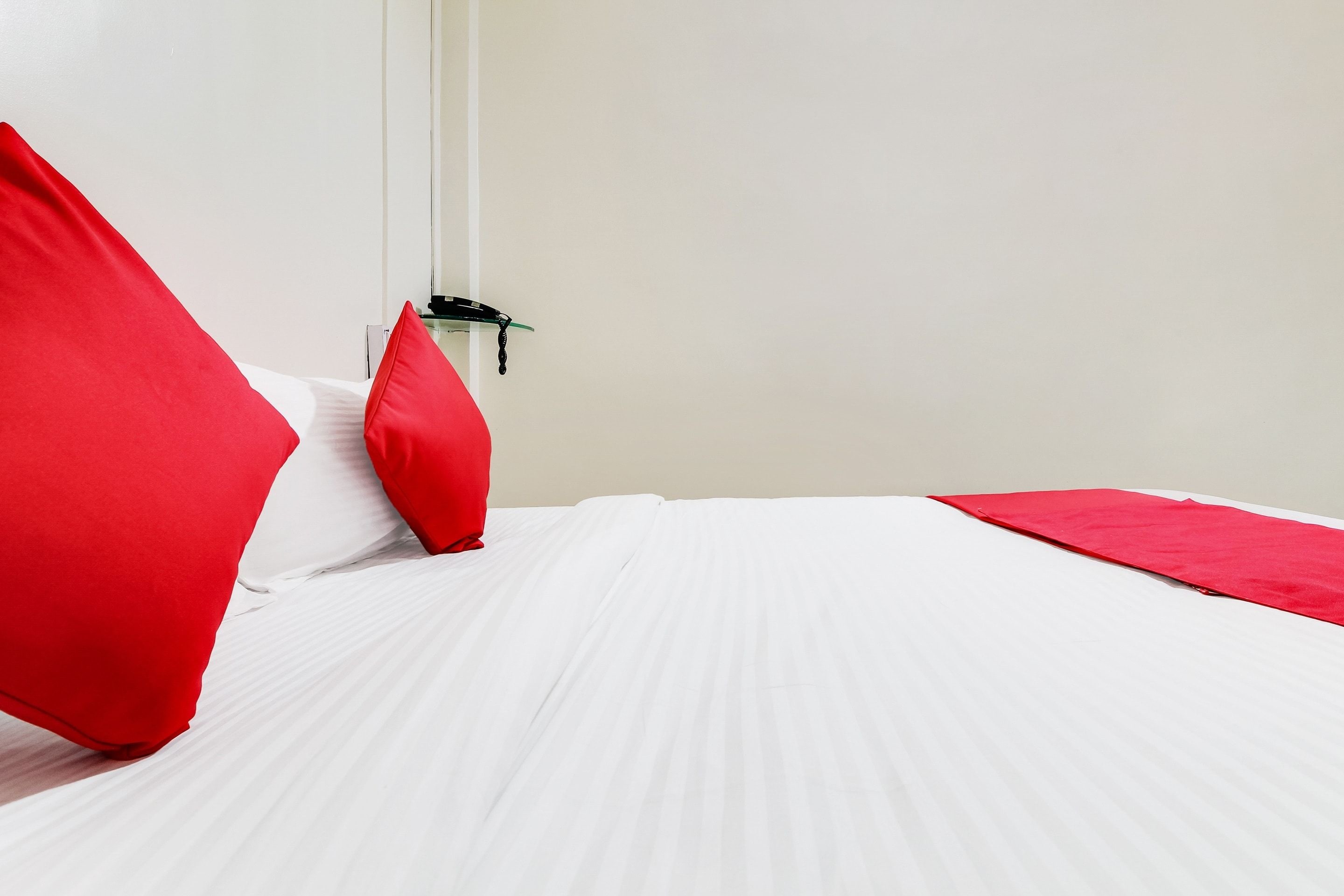 Uma cama com roupa de cama branca e almofadas vermelhas em um quarto com parede lisa e uma porta.
