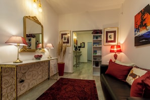 Uma sala de estar com tapete vermelho, sofá, mesa de consola com luminárias, espelho grande e um corredor que dá para uma cozinha.