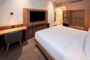 Quarto de hotel com cama, escrivaninha, cadeira, televisão na parede e guarda-roupa.