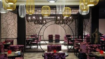 Restaurante com mesas redondas e cadeiras estofadas em roxo, com design geométrico na parede e luminárias pendentes.
