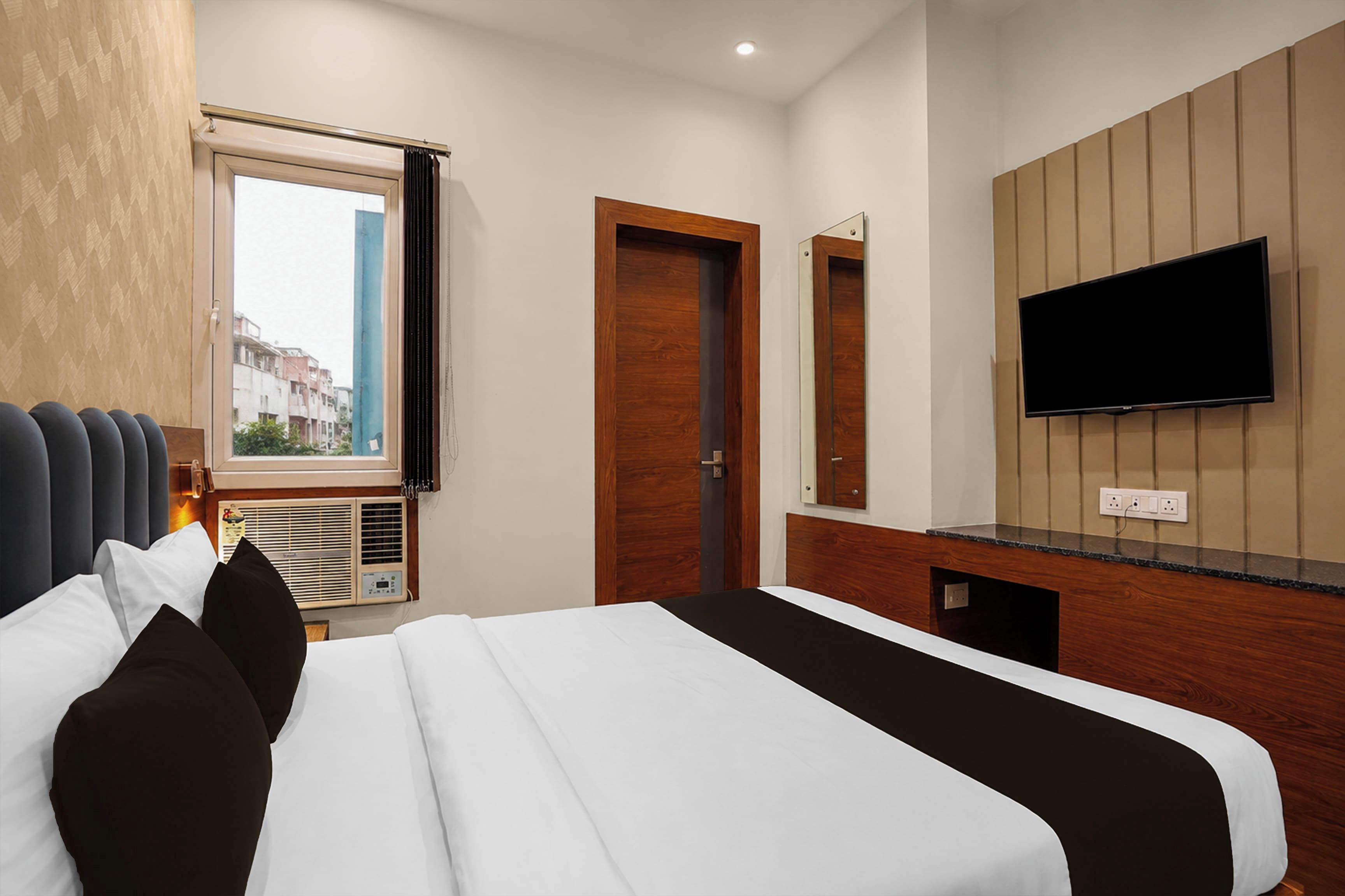 Quarto de hotel com cama, televisão fixada na parede, janela com cortinas e vista para prédios.