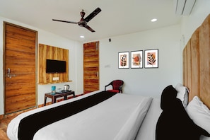 Quarto de hotel com cama, escrivaninha com chaleira, poltrona, ventoinha no teto e obras de arte na parede.