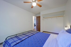 Quarto com cama, ventoinha no teto, porta e prateleira fixada na parede.