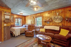 Interior rústico de cabana com cama, duas poltronas, um sofá, mesa de centro de madeira e ventilador de teto.