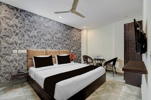 Quarto de hotel com cama, escrivaninha, cadeiras, ventoinha no teto e televisão.