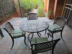 Pátio com mesa redonda e quatro cadeiras, cercado por um muro de tijolos e vegetação.