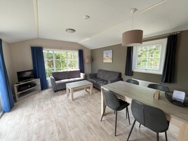 Uma sala de estar moderna com sofá, mesa de jantar e cadeiras, televisão em móvel e janelas amplas com cortinas.