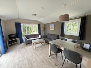 Uma sala de estar moderna com sofá, mesa de jantar e cadeiras, televisão em móvel e janelas amplas com cortinas.