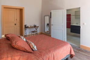 Quarto com cama, escrivaninha, cadeira, espelho e banheiro visível por uma porta aberta.