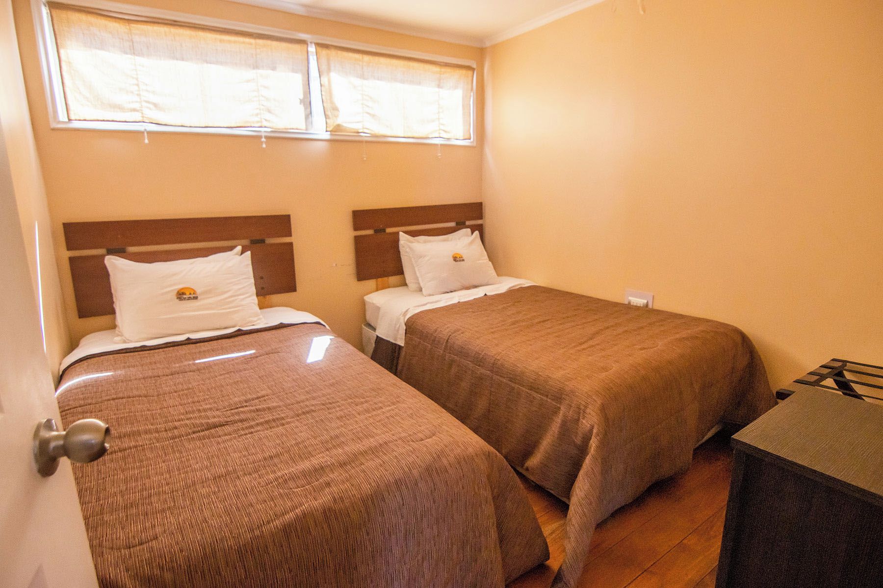 Quarto de hotel com duas camas de solteiro, uma mesinha e uma janela com persianas.