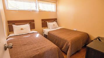 Quarto de hotel com duas camas de solteiro, uma mesinha e uma janela com persianas.
