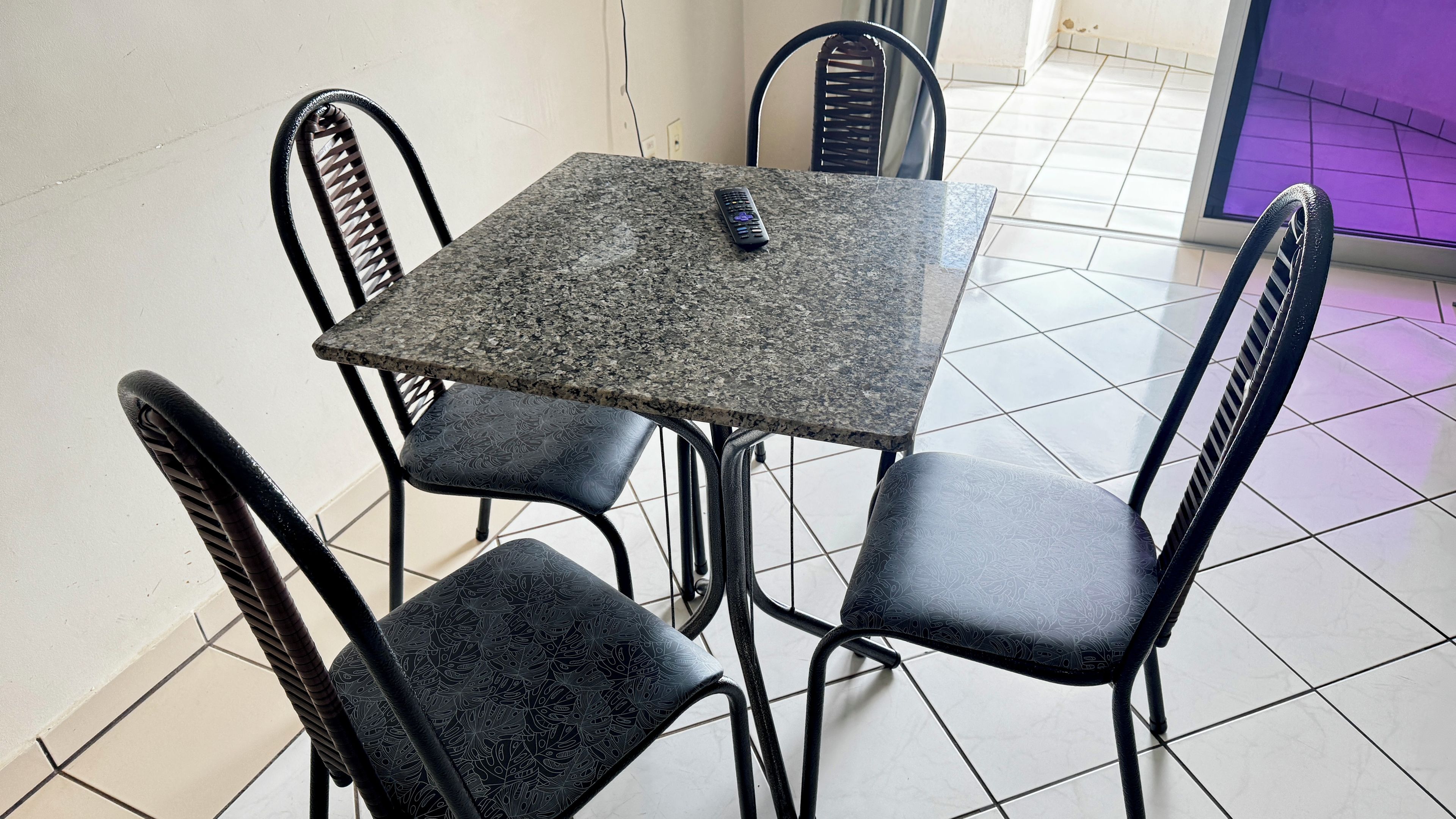 Mesa quadrada com controle remoto, rodeada por quatro cadeiras com padrão de folha.