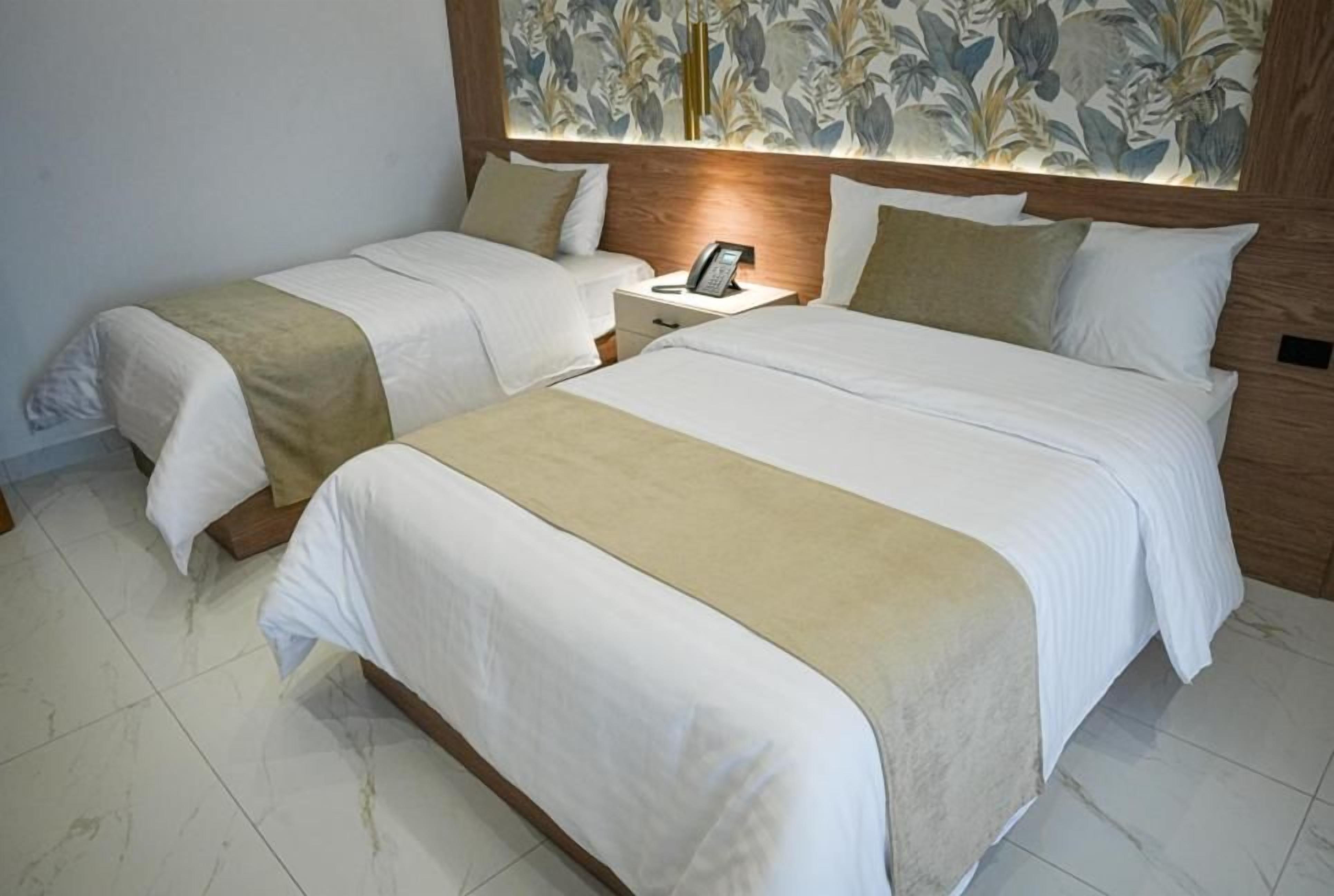 Quarto de hotel com duas camas, uma mesinha de cabeceira com telefone e papel de parede com motivos florais.